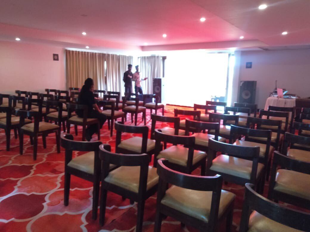 Um quarto com fileiras de cadeiras e mesas de madeira, um tapete com padrão vermelho e uma grande janela com cortinas.