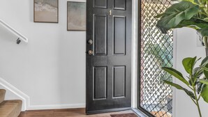 Uma porta preta com um número, uma parede branca com duas molduras, um chão de madeira e uma planta verde perto de uma porta de vidro.