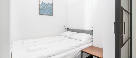 Uma cama de solteiro com roupa de cama branca, uma pequena mesa de madeira e um quadro na parede.