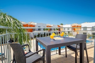 Uma varanda com mobiliário de vime, uma mesa com bebidas e uma piscina ao fundo.