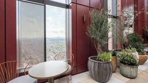 Varanda moderna com mesa redonda e cadeiras, plantas grandes em vasos e vista para a cidade através de grandes janelas.