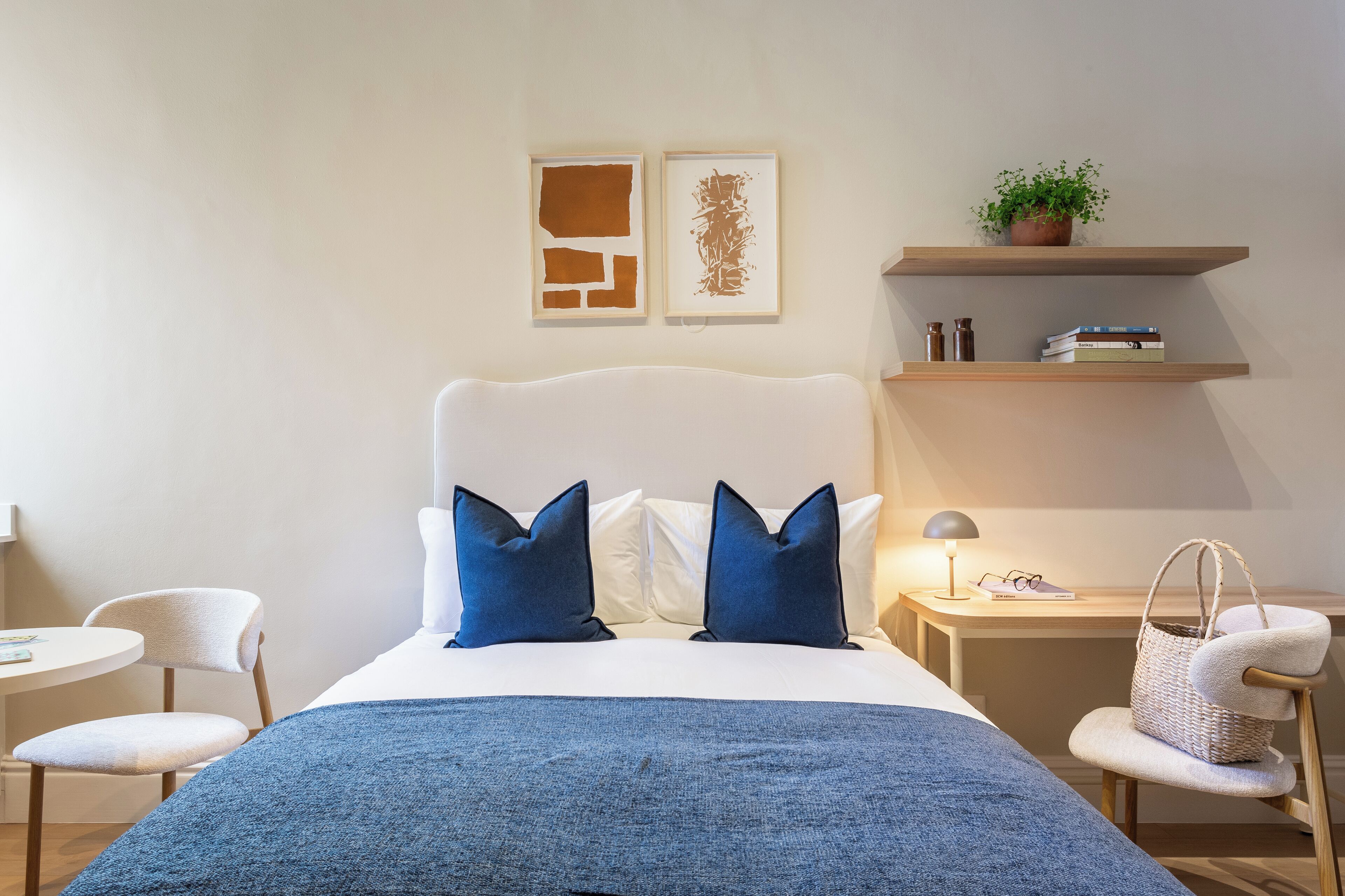 Quarto de hotel bem organizado, com uma cama, duas cadeiras, uma mesinha, um criado-mudo, uma luminária, uma planta e uma estante com livros e objetos decorativos.