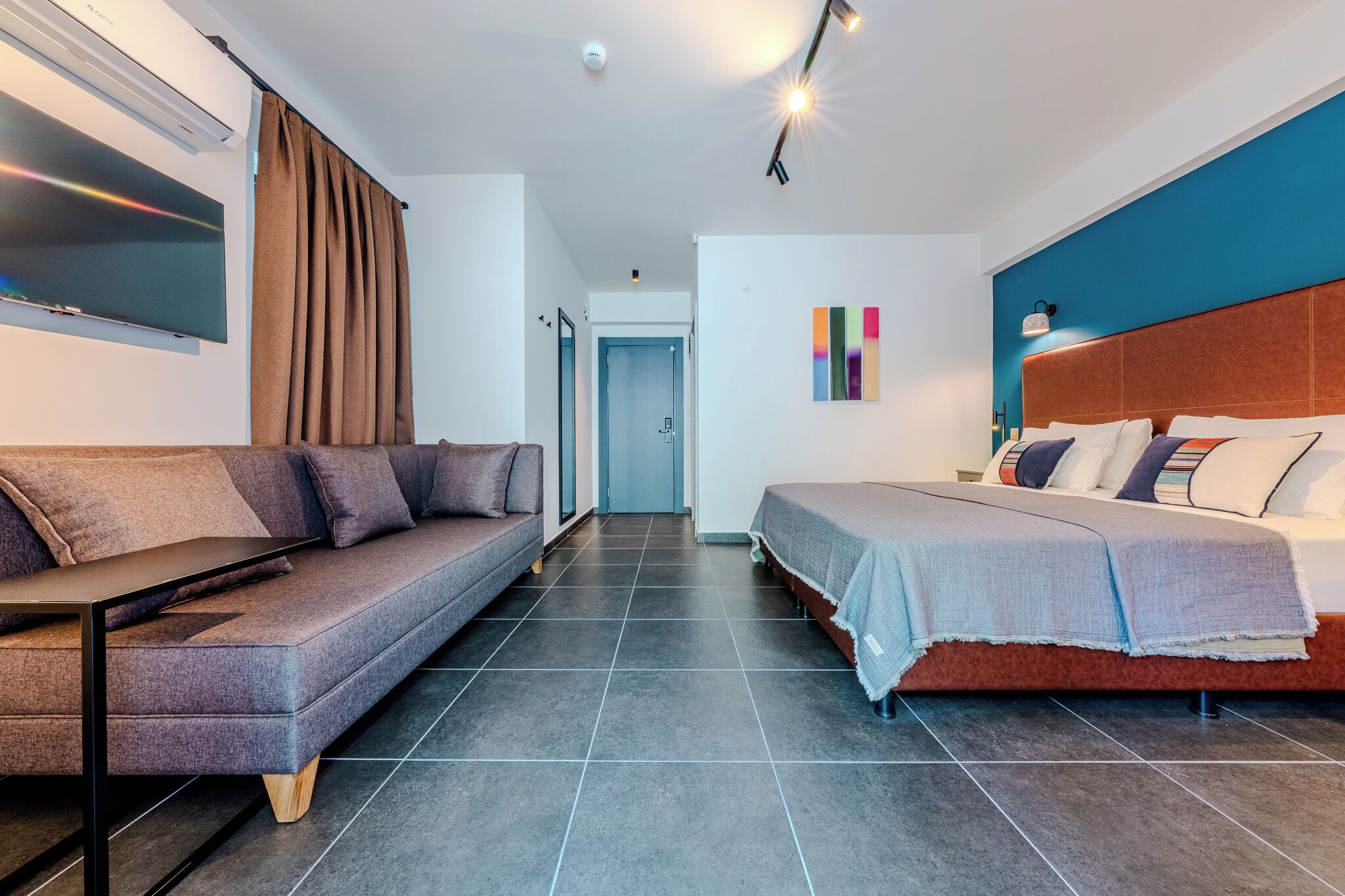 Quarto de hotel moderno com sofá, cama, televisão de tela plana e luminária embutida no teto.