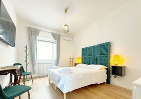 Quarto com duas camas, uma escrivaninha, cadeiras, uma janela com cortinas e uma luminária na parede.