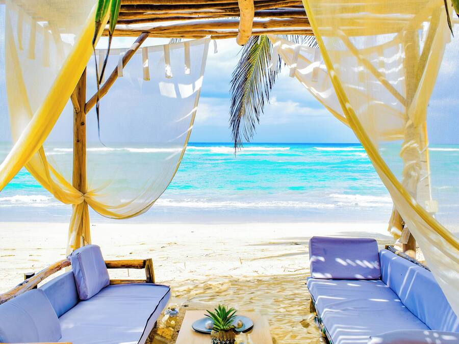 Uma cabana de praia com duas áreas de assento acolchoadas, uma mesa com uma travessa de frutas e vista para o oceano.
