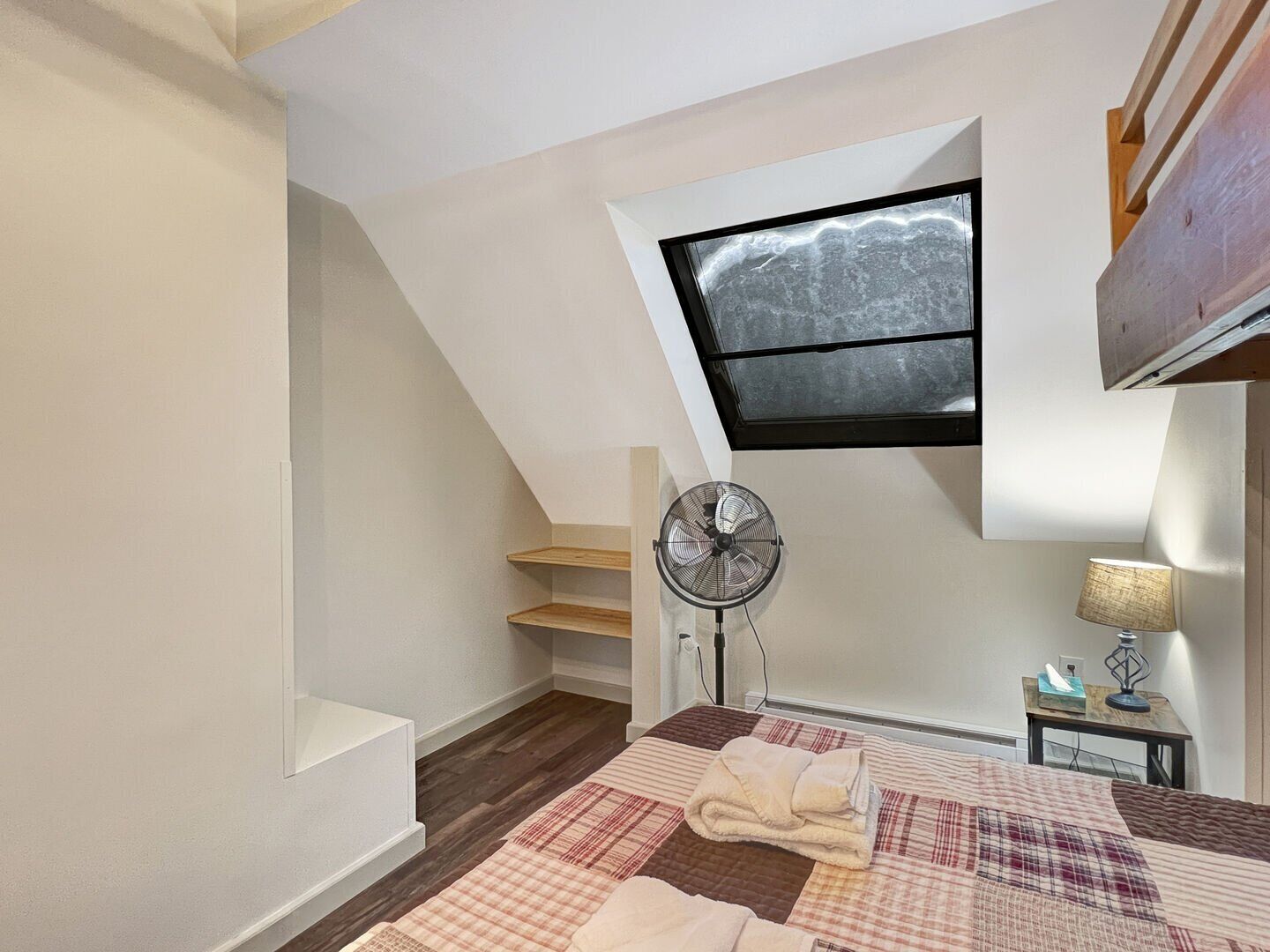 Um quarto com teto inclinado, um claraboia, uma cama com colcha xadrez, um ventilador e uma mesa de cabeceira com abajur.