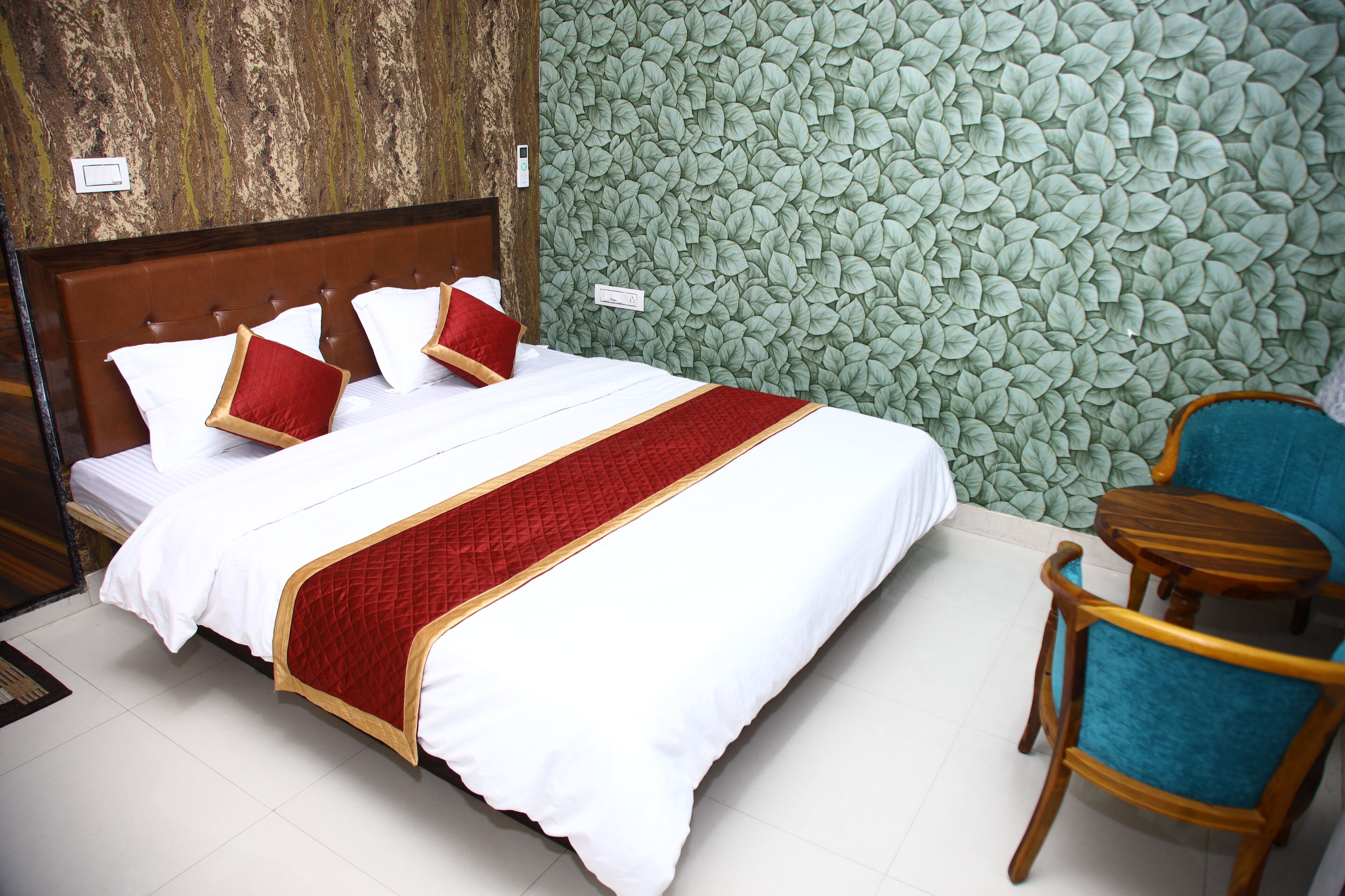 Cama bem arrumada com roupa de cama branca e detalhes vermelhos, cabeceira de madeira e parede com padrão de folhas verdes. Há uma cadeira de madeira com almofada azul à direita.