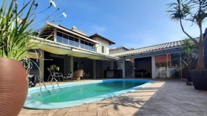 Uma piscina com deck azulejado, um alpendre coberto e uma casa de dois andares com telhado em azulejos.