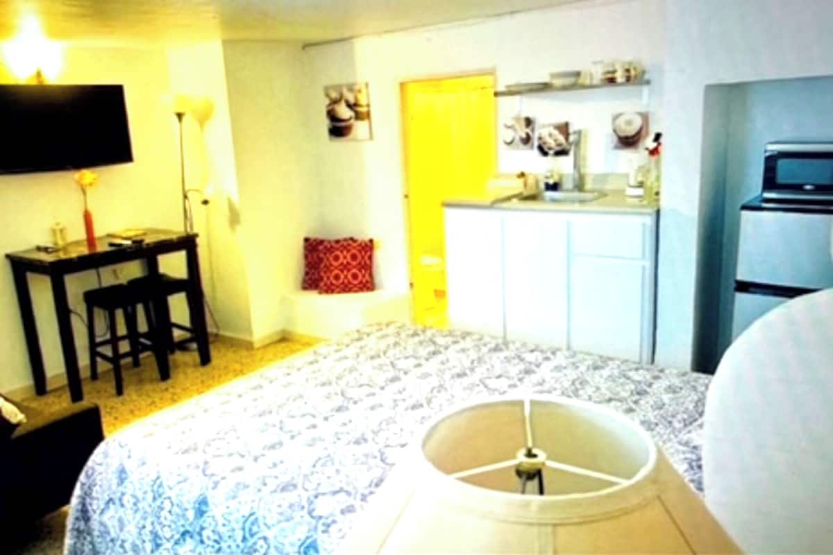 Quarto com cama, escrivaninha e cadeira. Possui área de cozinha com geladeira e micro-ondas.