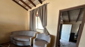 Um quarto com sofá curvo e uma cadeira, janela com cortinas e porta que dá para outro cômodo.