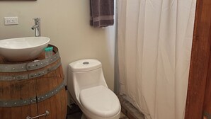 Casa de banho com lavatório em barrica de madeira, sanita branca, suporte de toalhas com toalha escura e cortina de chuveiro.