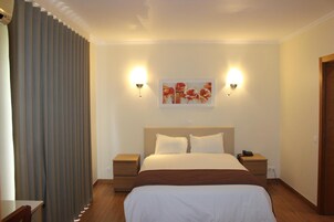 Quarto de hotel com cama, duas mesas de cabeceira, escrivaninha, cadeira, quadro na parede e janela com cortinas.