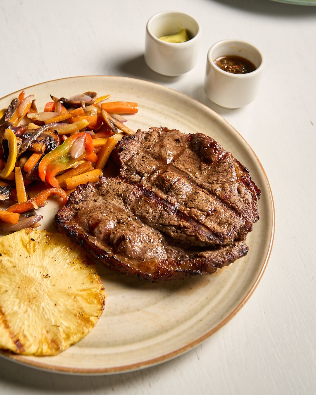 Um prato com bife grelhado, legumes salteados e uma fatia de abacaxi.