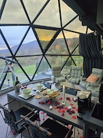 Uma sala de jantar com uma mesa posta para dois, um telescópio e uma vista panorâmica através de uma grande janela.