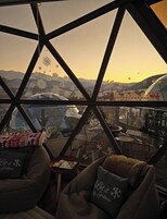 Quarto aconchegante com vista para montanhas cobertas de neve ao pôr do sol, apresentando uma estrutura de vidro geométrica, uma mesa pequena com um objeto decorativo e um sofá com uma almofada.