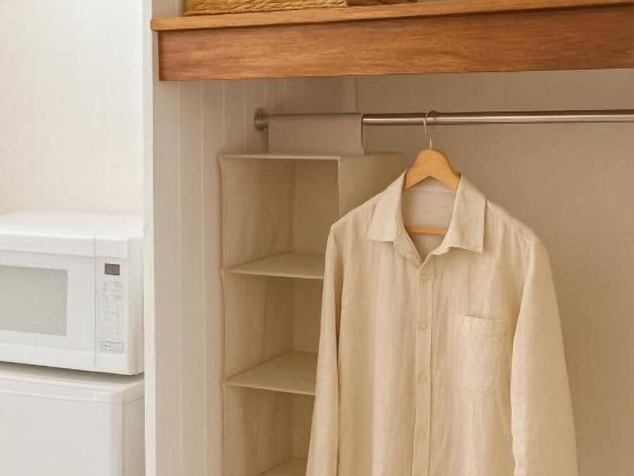 Um micro-ondas branco sobre um armário branco, uma prateleira de madeira com uma cesta de palha, e uma camisa branca pendurada num guarda-roupa com prateleiras abertas.