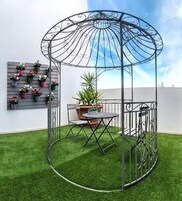 Um gazebo de metal com cobertura de vidro, cercado por grama sintética e um vaso de planta no centro.