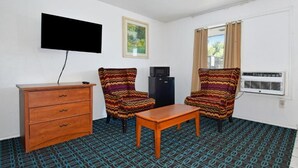 Um quarto pequeno com cômoda de madeira, duas poltronas estampadas, uma mesa pequena, uma TV de tela plana, um quadro emoldurado e um aparelho de ar condicionado.