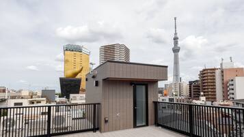Um edifício moderno com porta de vidro e guarda-corpo de metal, cercado por um horizonte urbano com diversos edifícios e uma torre.