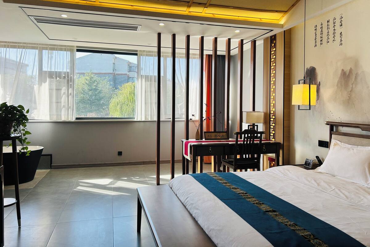 Quarto de hotel moderno com cama, escrivaninha e cadeira, uma planta em vaso e vista para prédios.
