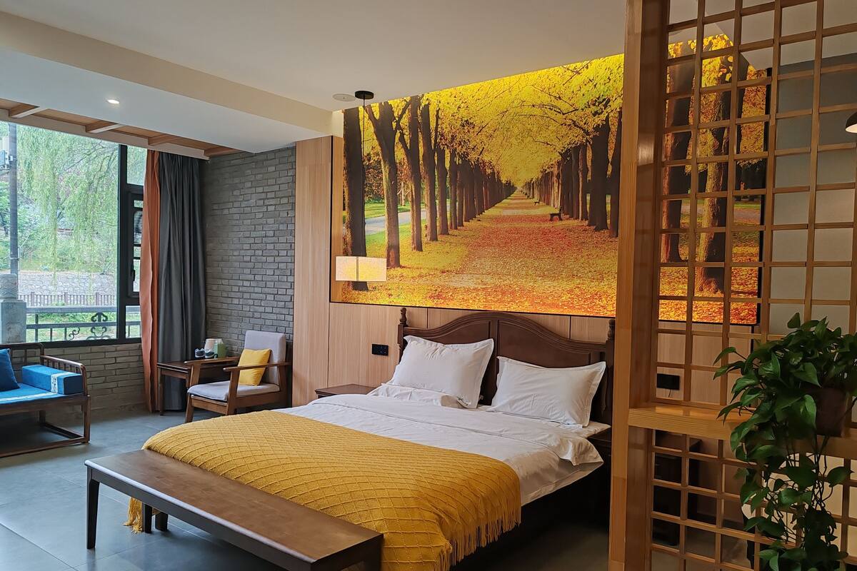 Quarto de hotel moderno com uma cama grande, um banco, uma cadeira, uma mesinha de cabeceira e uma janela ampla com vista para árvores.