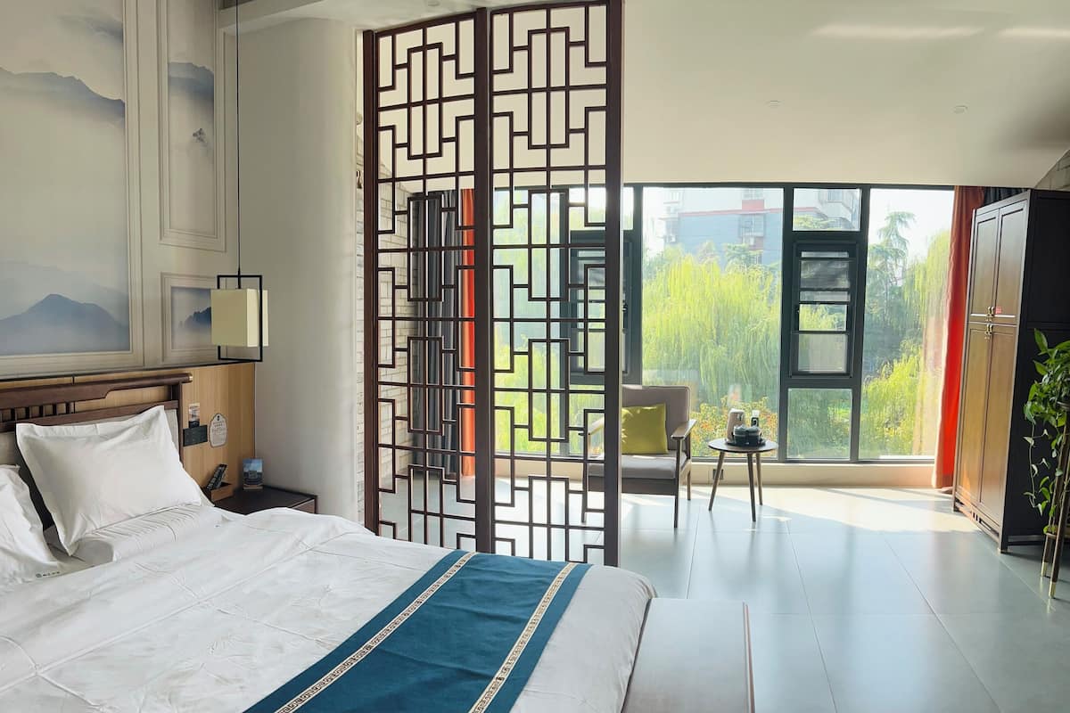 Quarto de hotel com uma cama grande, vista para o verde através de uma porta deslizante, uma mesinha com uma cadeira e uma mesa de cabeceira com abajur.