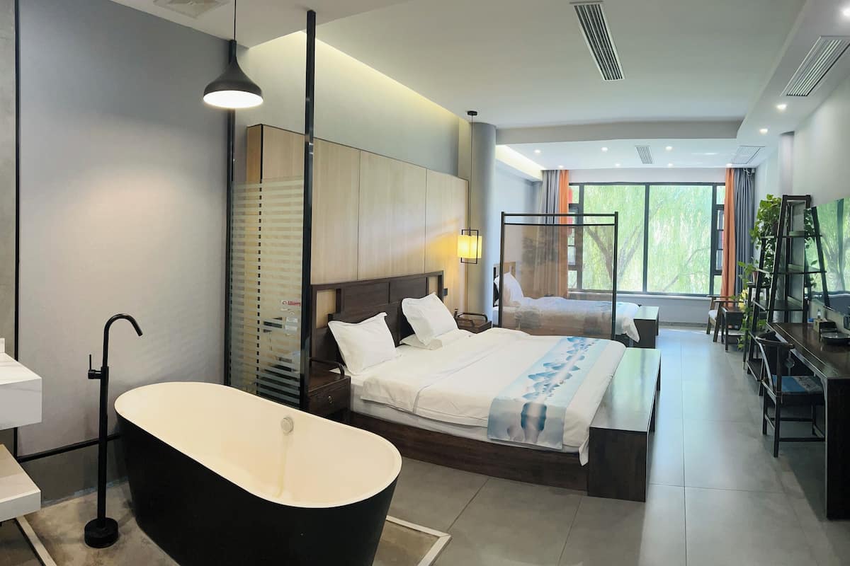 Quarto de hotel moderno com uma cama grande, uma banheira, uma escrivaninha e uma janela ampla com vista para árvores.