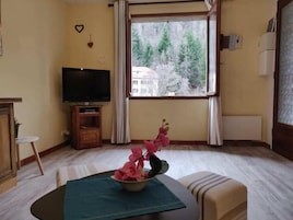 Uma sala de estar aconchegante com televisão, uma mesinha com um vaso de flores e uma janela grande com vista para uma floresta.