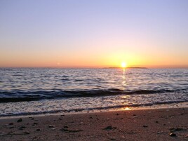 Uma praia com o sol se pondo sobre a água, ondas calmas e uma faixa de areia com pedras espalhadas.