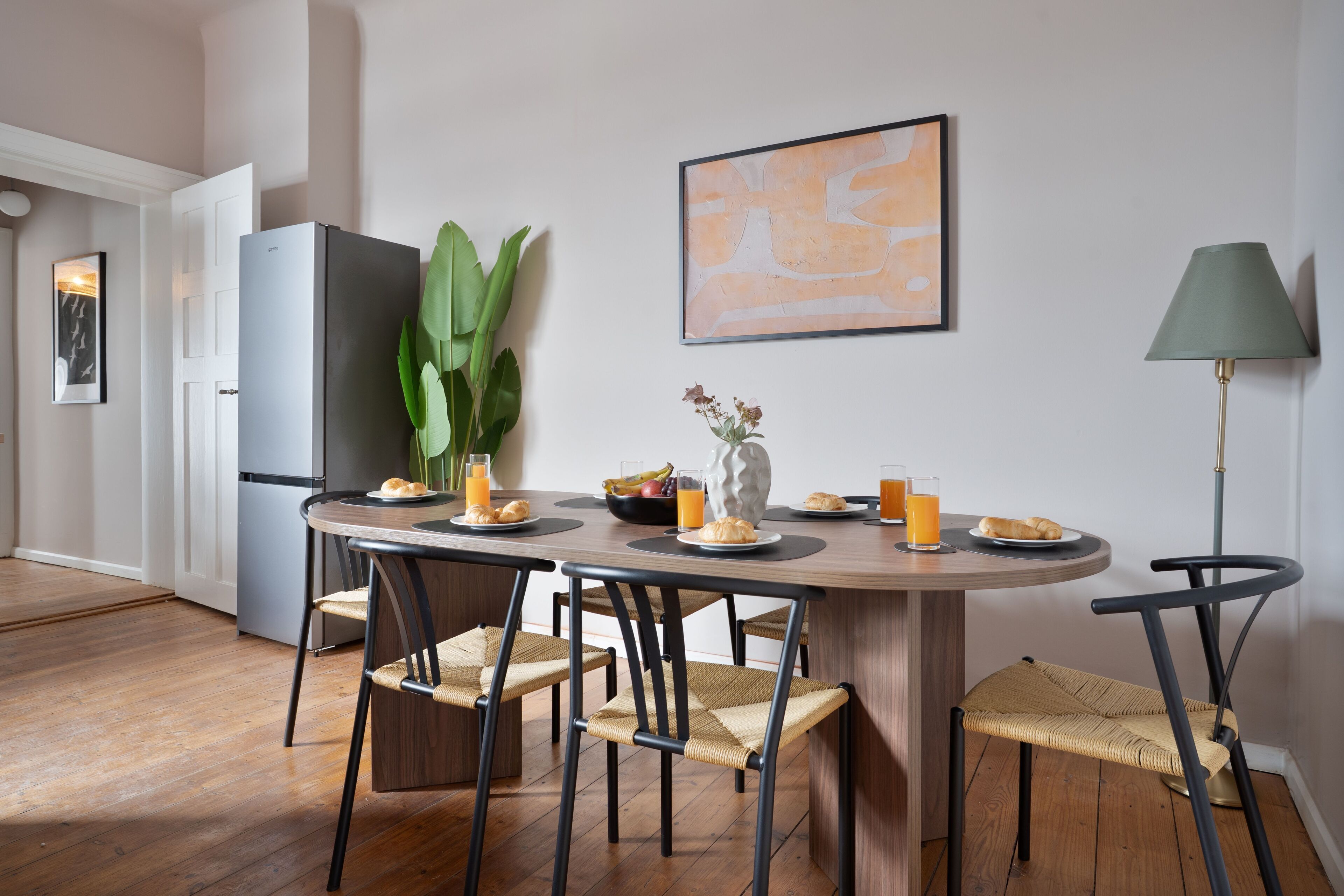 Uma área de refeições com mesa de madeira, quatro cadeiras, geladeira, planta em vaso, vaso com flores e um abajur.