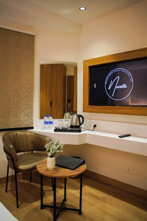 Quarto de hotel com escrivaninha de madeira, uma cadeira, uma mesinha com um vaso, televisão na parede e um espelho.