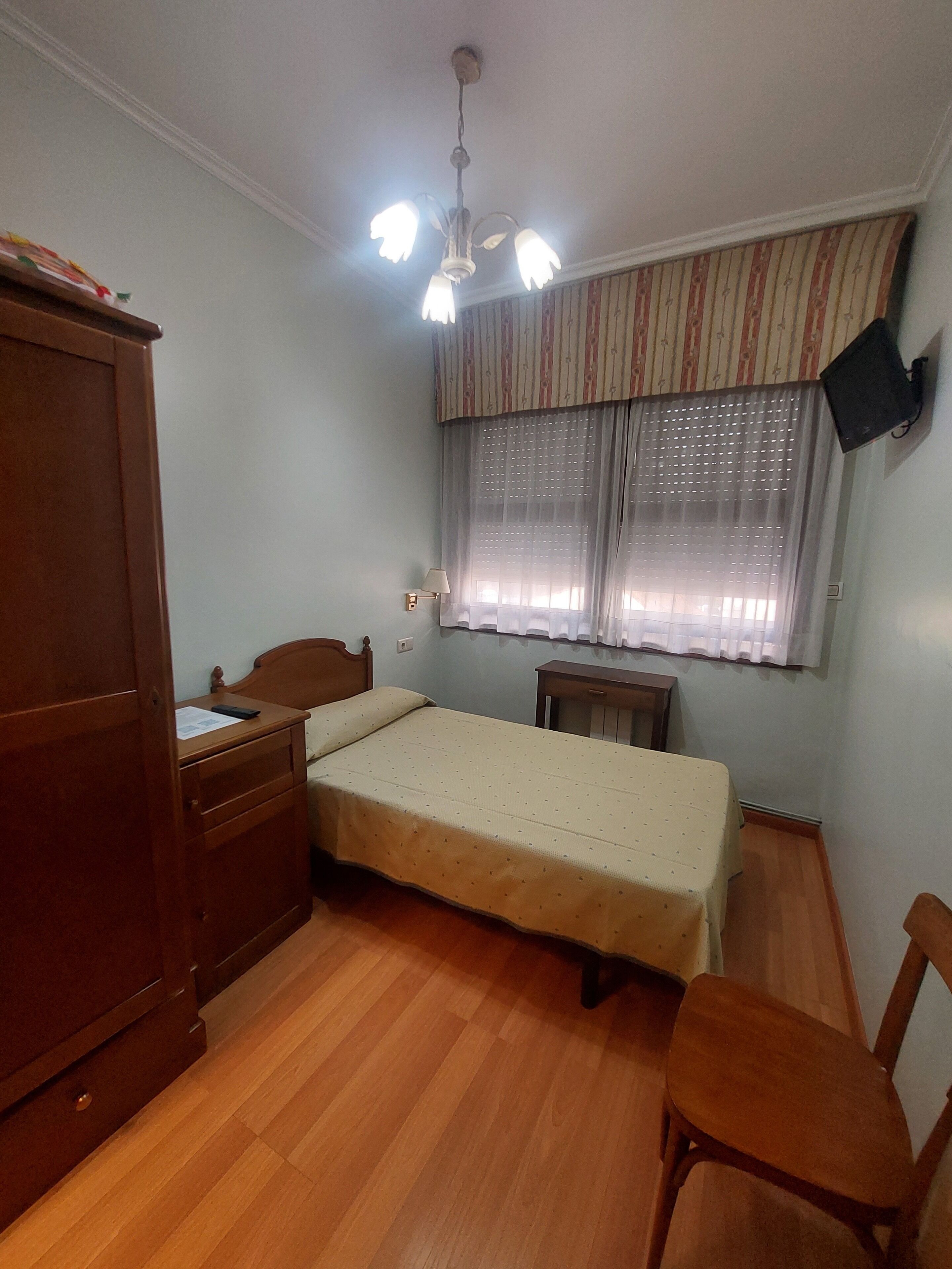 Quarto com cama, guarda-roupa de madeira, escrivaninha, cadeira e janela com cortinas.