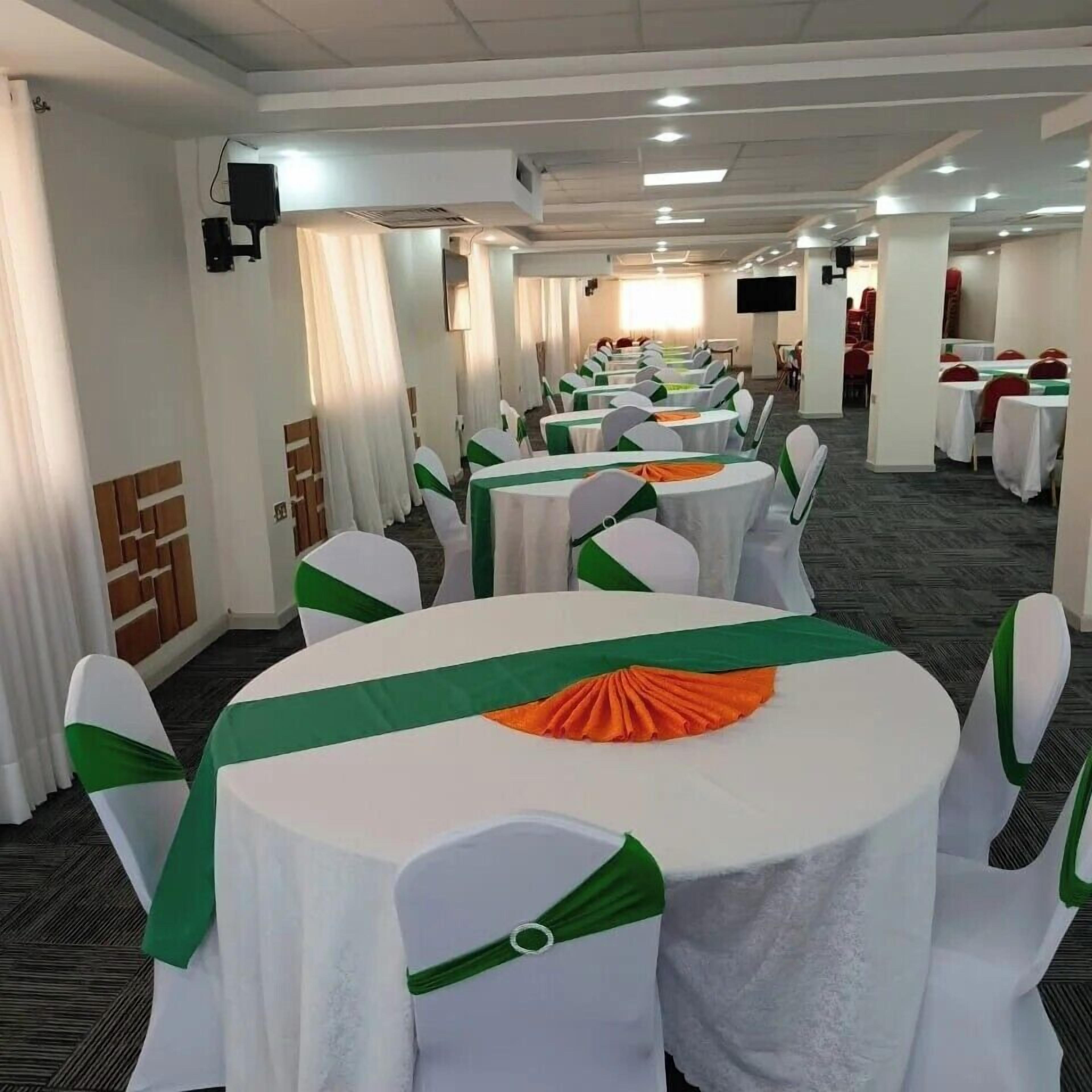 Uma sala de banquetes com mesas redondas, toalhas brancas, fitas verdes e centros de mesa laranja.