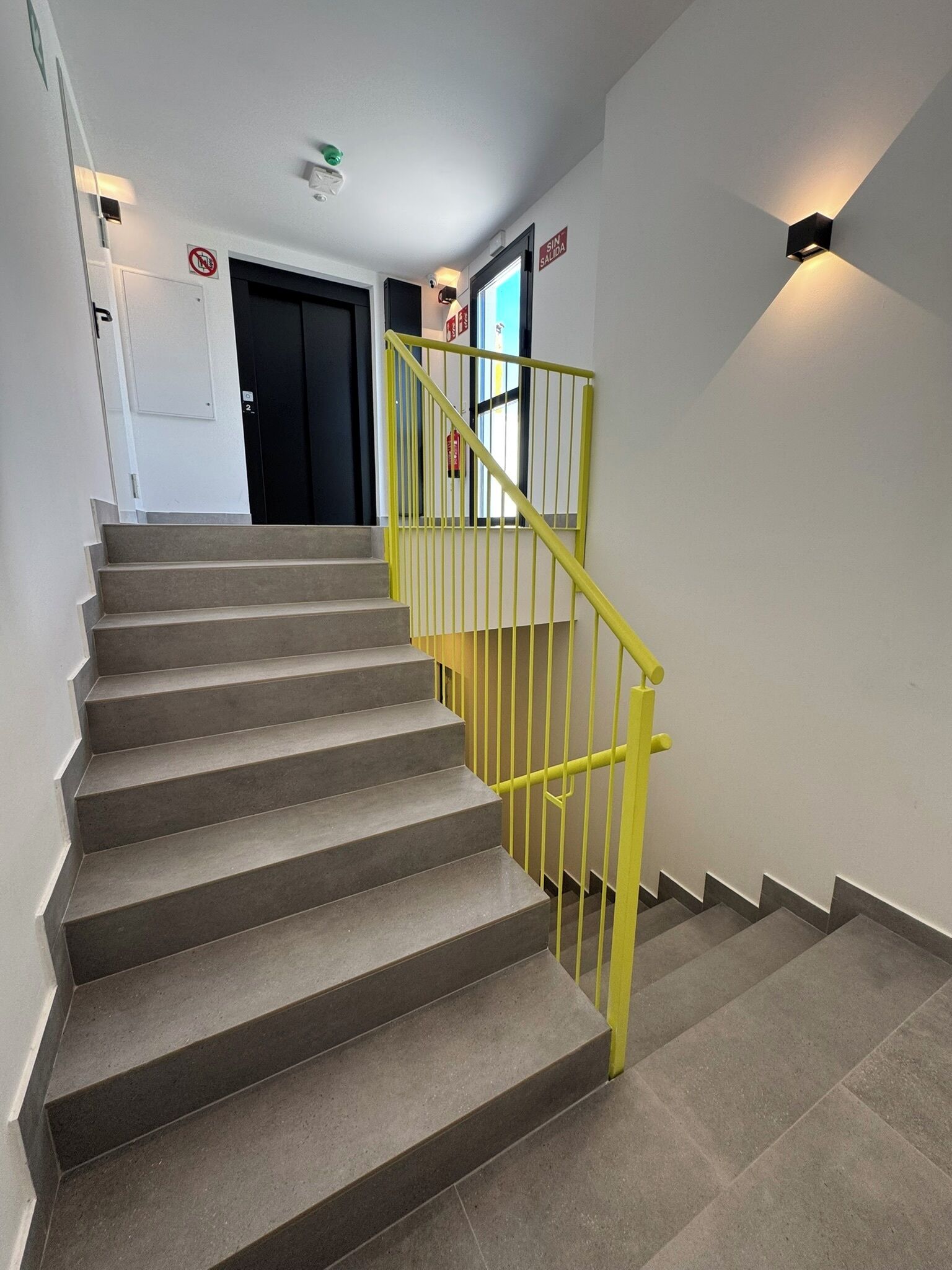 Uma escadaria com degraus cinzentos e corrimãos amarelos que levam a uma porta preta.