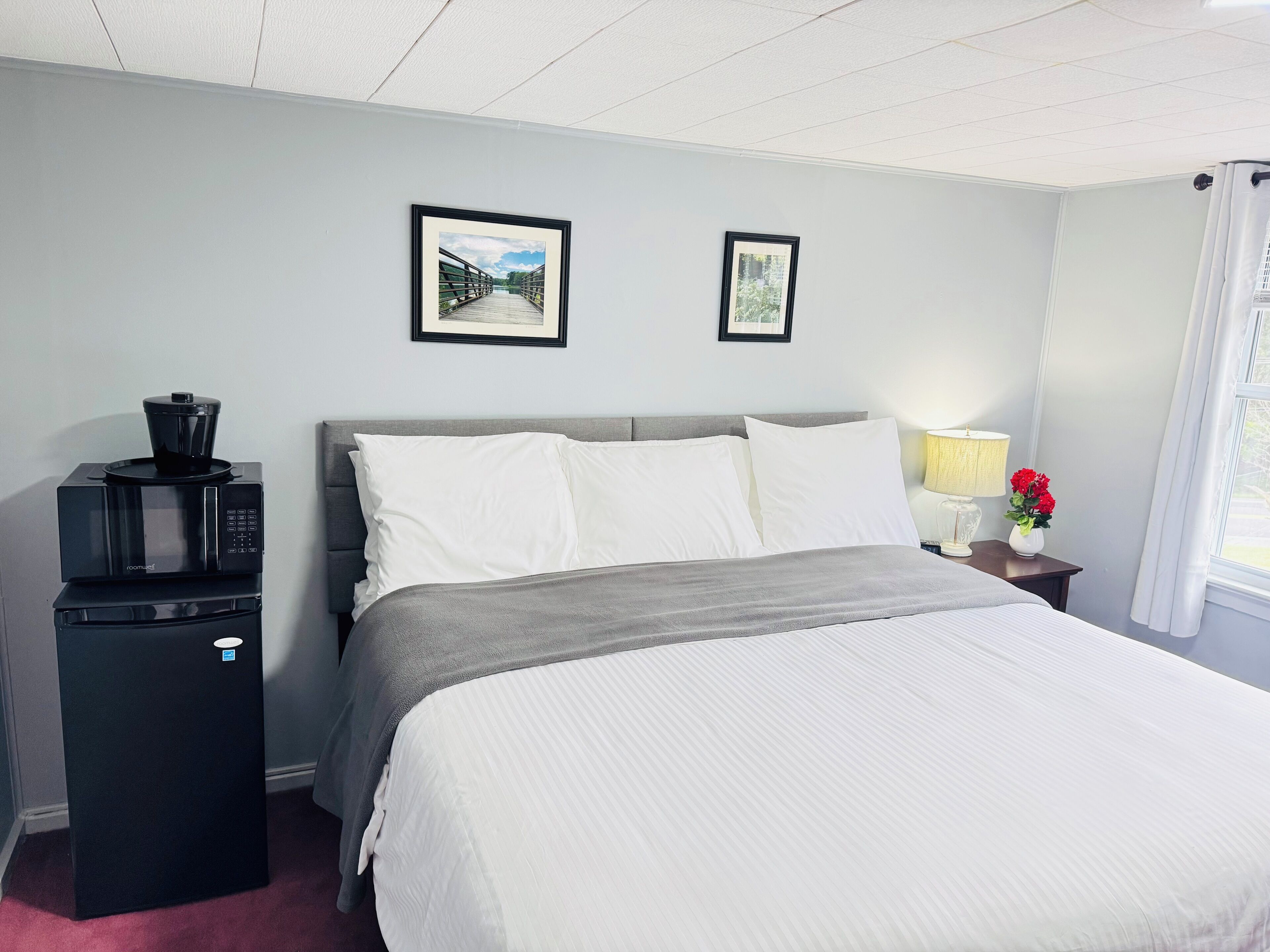 Quarto de hotel com uma cama, cafeteira, mesa de cabeceira com abajur e duas fotos emolduradas na parede.