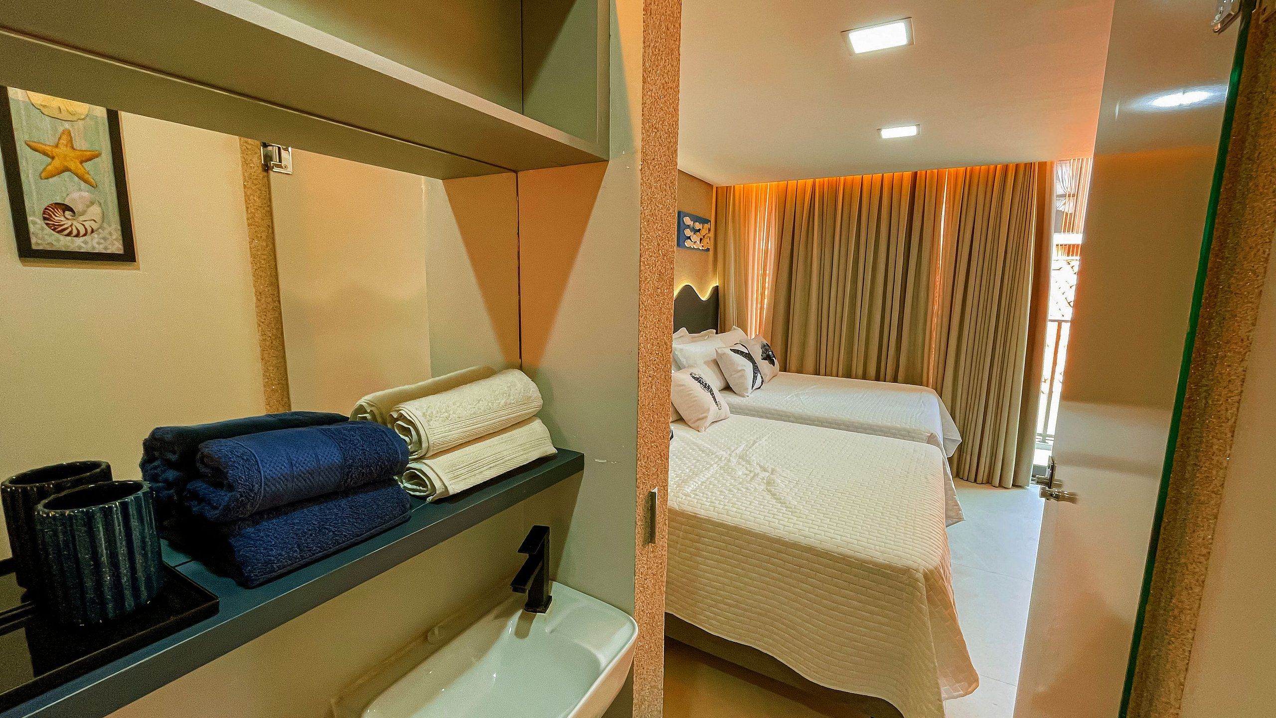 Quarto de hotel com cama, pia, espelho e prateleira com toalhas e caneca.