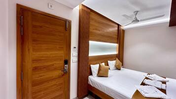 Quarto de hotel com porta de madeira, cama com roupa de cama branca e duas almofadas, ventoinha no teto e janela com vidro fosco.