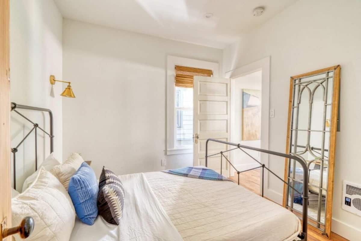 Quarto com estrutura de cama de metal, mesa de cabeceira, luminária e janela com persianas.