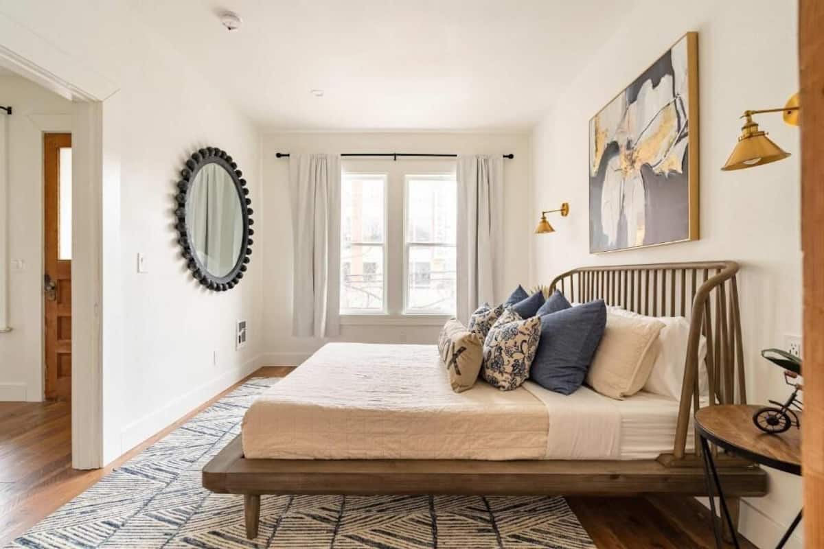 Quarto com cama, espelho redondo, janela com cortinas, um quadro e uma luminária.