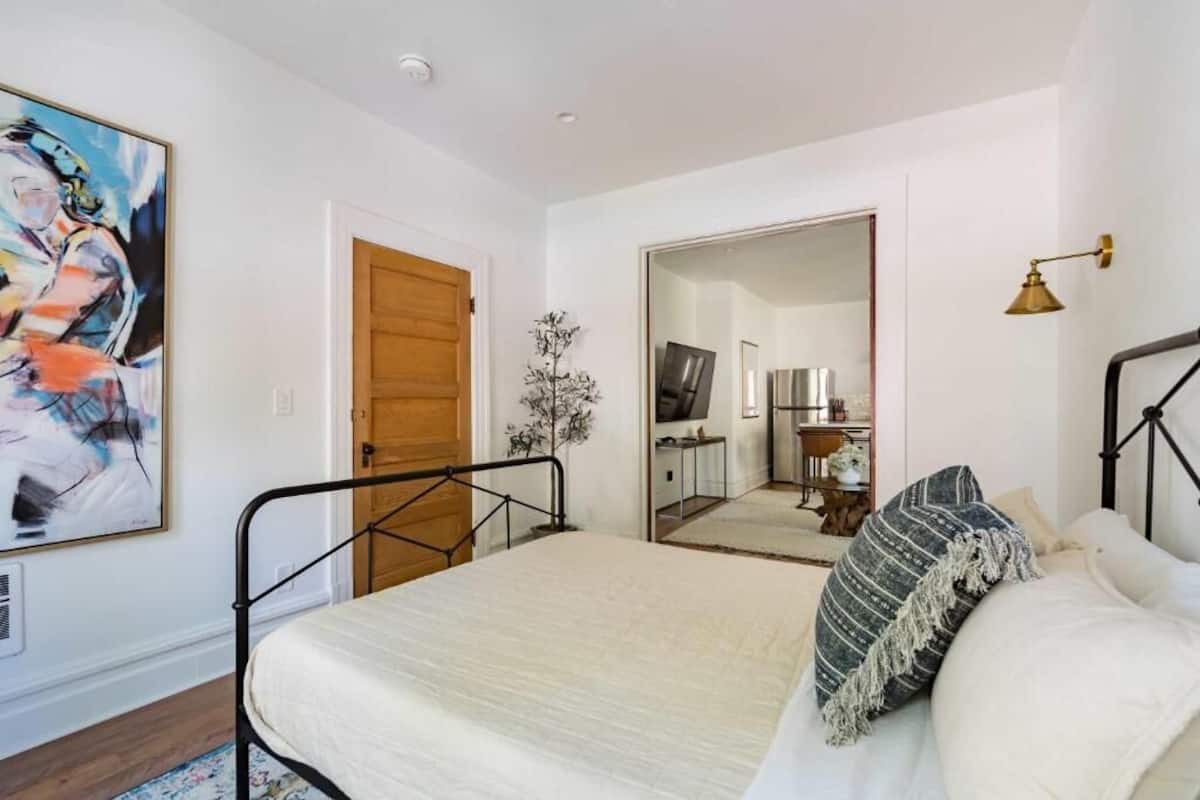 Quarto com cama, porta de madeira, espelho, uma pequena planta e um quadro na parede.
