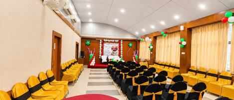 Uma sala de conferências com fileiras de cadeiras amarelas e pretas, uma mesa vermelha e um palco com um banner.