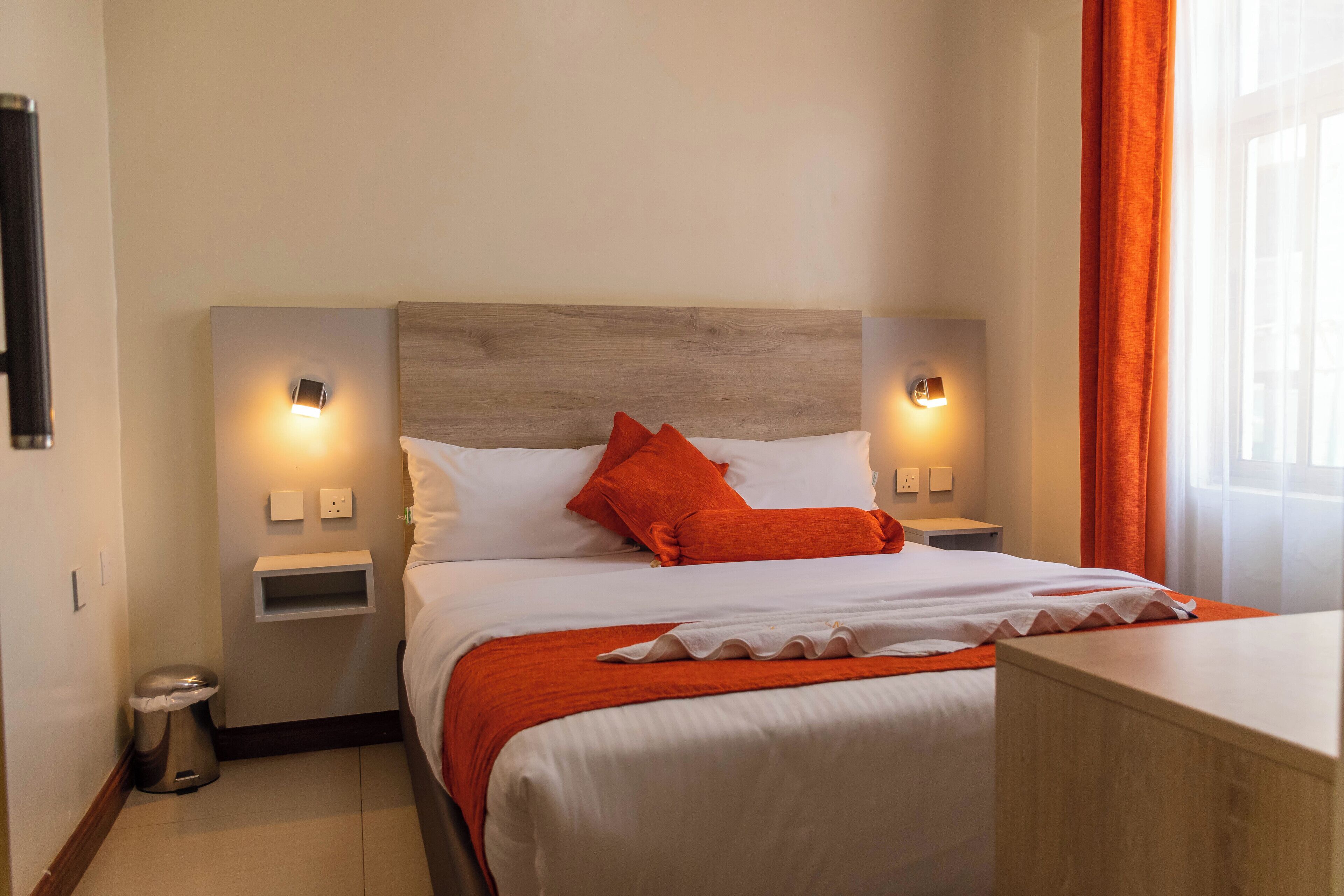 Cama bem arrumada com roupa de cama branca e laranja, duas mesas de cabeceira com abajures, uma pequena lixeira e uma janela com cortinas laranja.