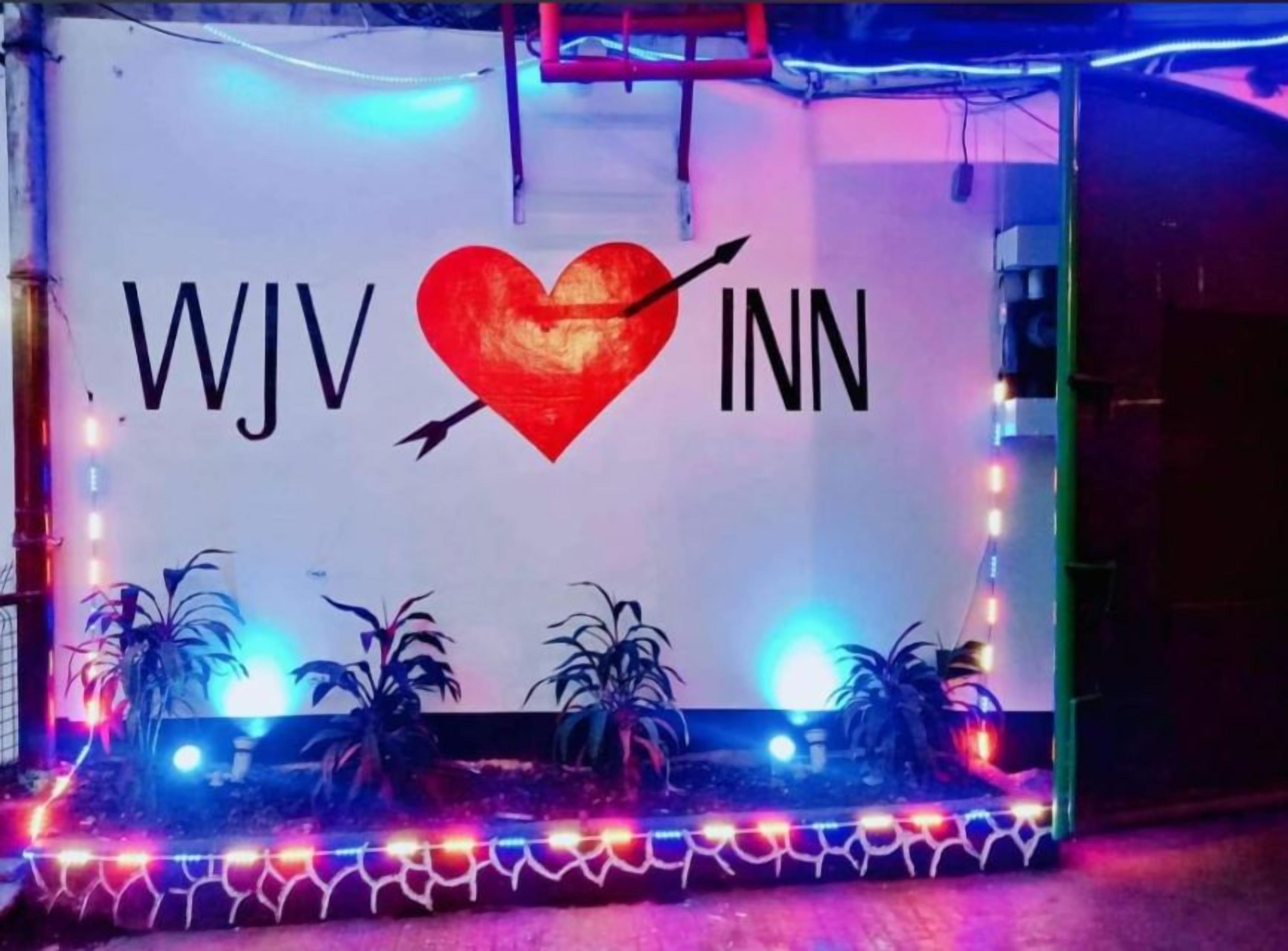 Um letreiro com o texto 'WJV INN' e um símbolo de coração, rodeado por luzes decorativas e plantas em vasos.