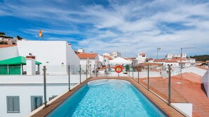 Piscina com vista para uma cidade costeira, cercada por edifícios com telhados de barro vermelhos.