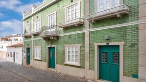 Uma fileira de edifícios verdes com molduras de janelas e varandas brancas, uma rua de paralelepípedos e um céu azul límpido.