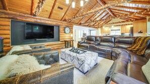 Interior de cabana de tronco com uma televisão grande, sofás de couro e uma lareira acolhedora.