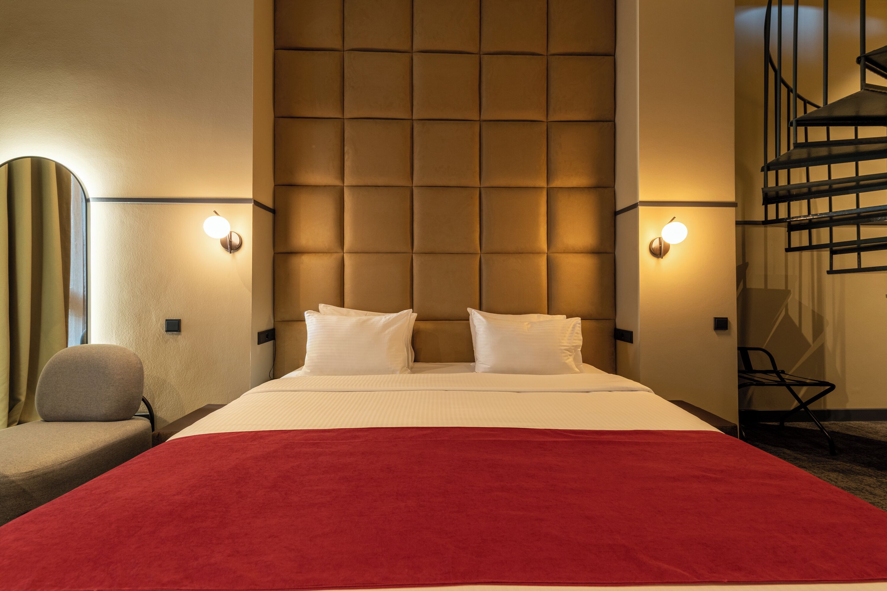 Quarto de hotel com uma cama grande, cabeceira acolchoada, duas luminárias de cabeceira, uma cadeira e uma escada.
