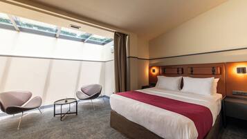 Quarto de hotel com uma cama grande, duas poltronas, uma mesinha e uma janela com vista.