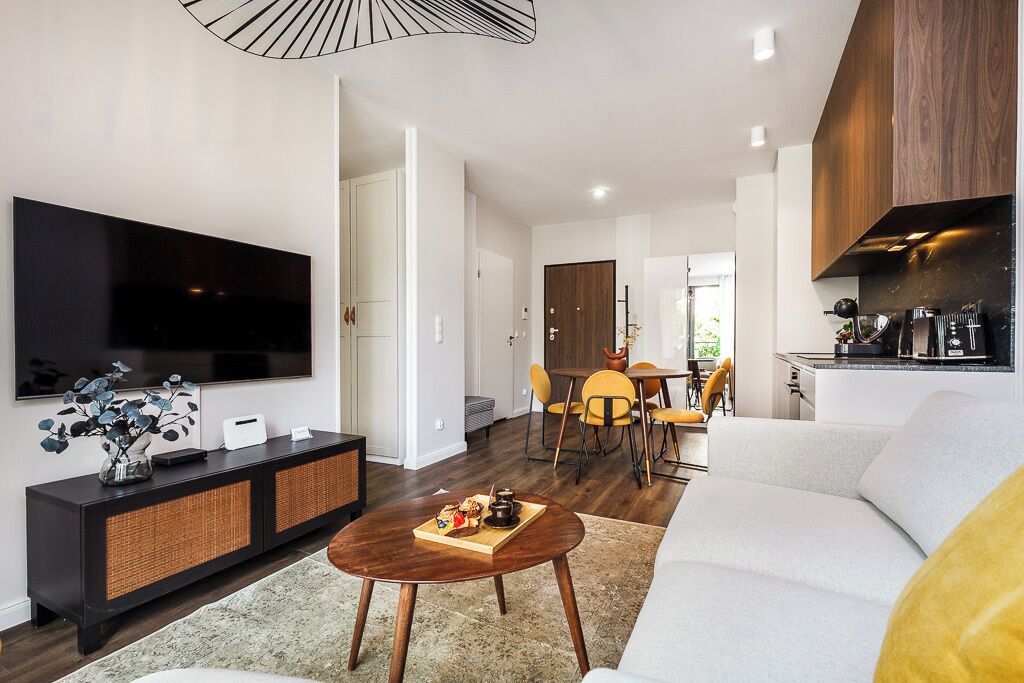Uma sala de estar moderna com televisão de tela plana, uma mesa de centro de madeira com uma bandeja de petiscos, um sofá e uma área de jantar com cadeiras amarelas.