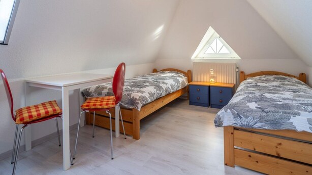 Um quarto de sótão pequeno e triangular com duas camas de solteiro, uma escrivaninha e uma cadeira.